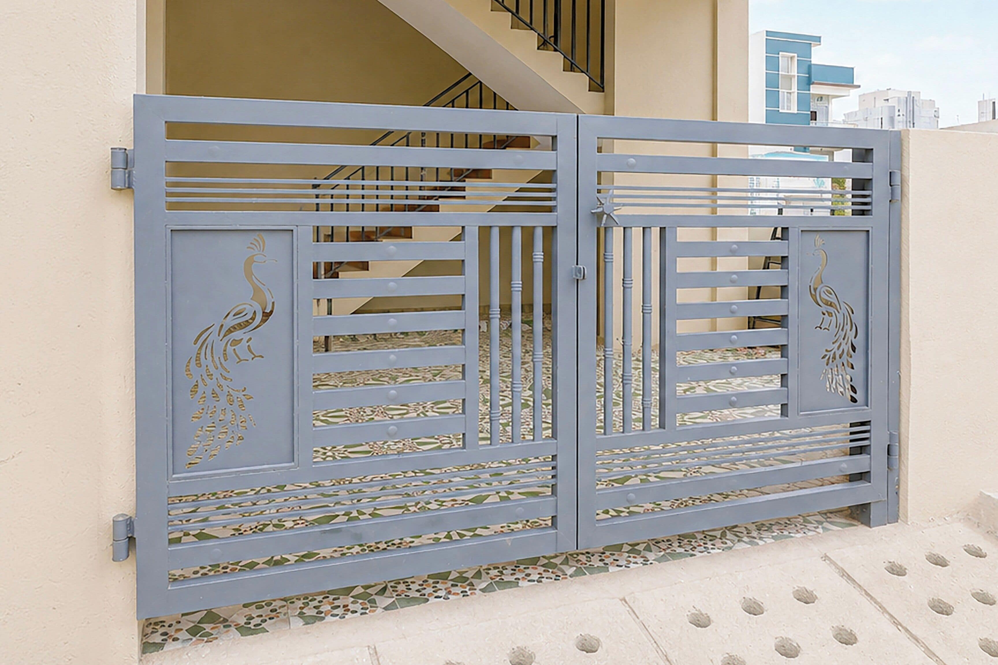 Um portão de metal cinzento com detalhes decorativos e um desenho de pavão, dando acesso a uma escadaria.