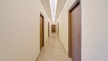 Um corredor longo e estreito com portas em ambos os lados, que dá acesso a quartos. O chão é ladrilhado e as paredes têm uma cor clara.