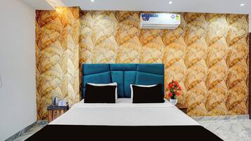 Quarto de hotel com uma cama grande, cabeceira azul, ar condicionado fixado na parede e um painel decorativo com padrão de folhas.