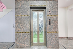 Um elevador com porta de vidro, inserido em um edifício com parede de pedra estampada e moldura de porta branca.