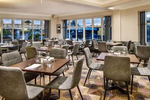 Espaço de refeições de hotel com várias mesas e cadeiras, grandes janelas e um tapete estampado.