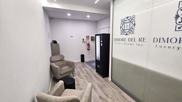Corredor com poltrona de massagem, extintor de incêndio e parede com logotipo e texto para 'DIMORE DEL RE Luxury Rooms Spa'.