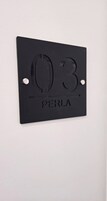 Placa de metal preta com os números '03' e a palavra 'PERLA' gravados, fixada em uma parede branca.