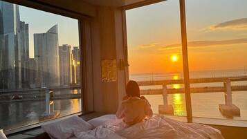 Quarto de hotel com grande janela vista para paisagem urbana e pôr do sol.