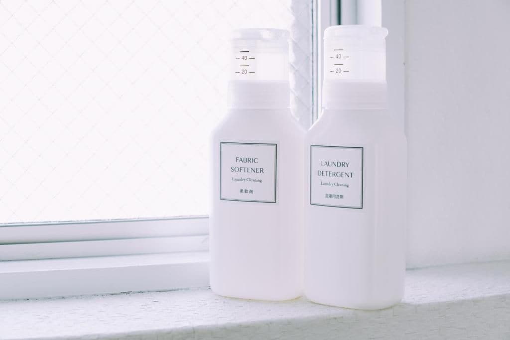 Duas garrafas brancas, uma com a etiqueta 'FABRIC SOFTENER' e outra com a etiqueta 'LAUNDRY DETERGENT', sobre uma superfície branca perto de uma janela.
