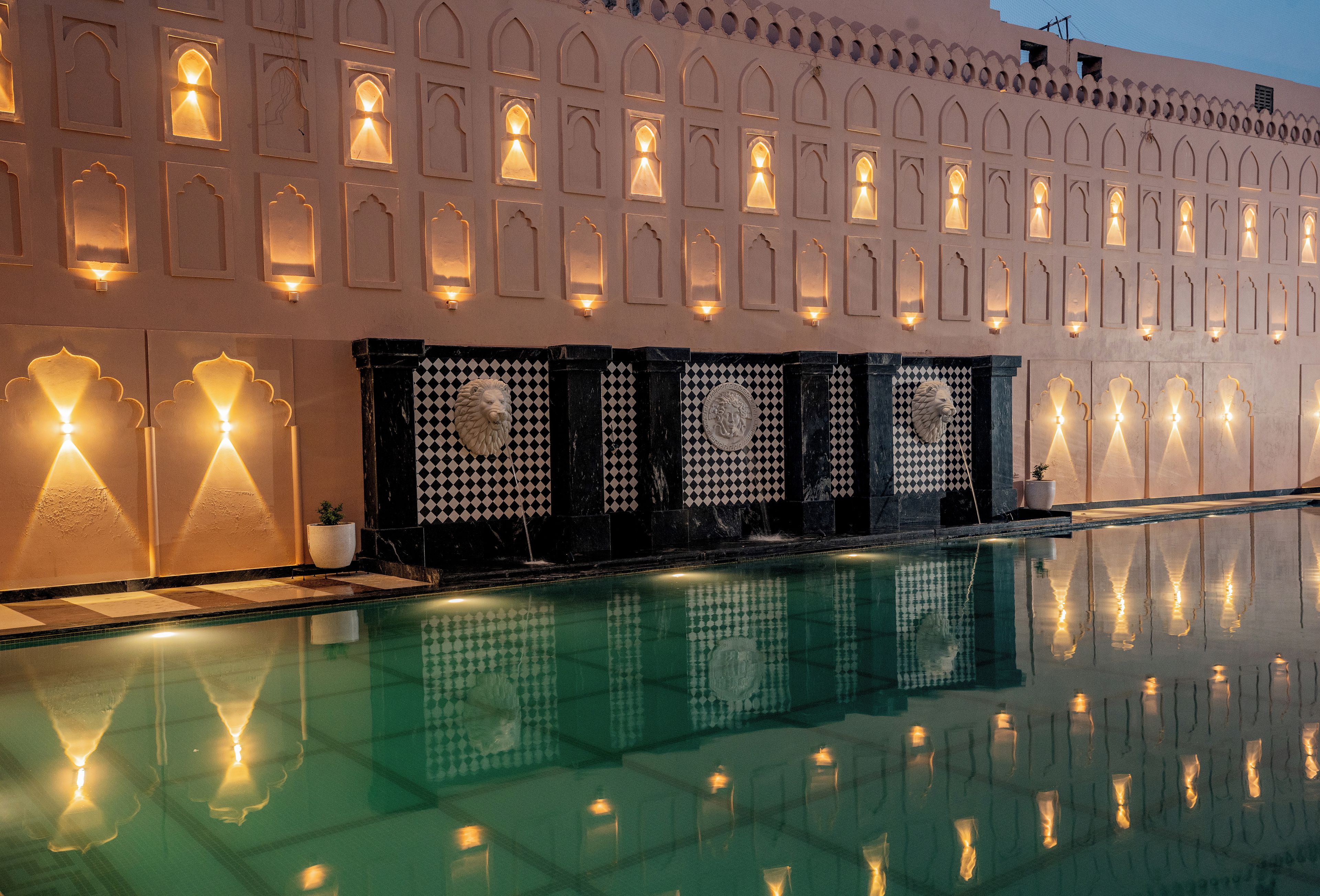 Piscina com reflexo da fachada do edifício, que inclui uma série de janelas em arco e painéis decorativos na parede.
