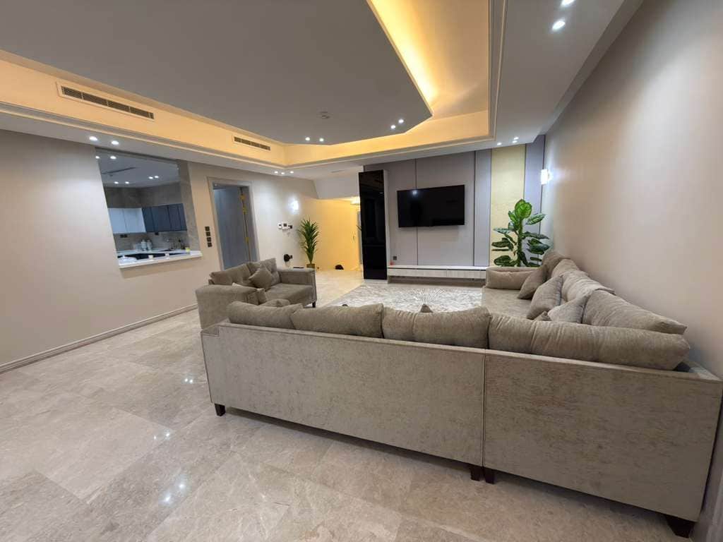 Uma sala de estar moderna com um sofá seccional, uma TV de tela plana na parede e uma cozinha visível através de uma porta aberta.