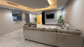 Uma sala de estar moderna com um sofá seccional, uma TV de tela plana na parede e uma cozinha visível através de uma porta aberta.