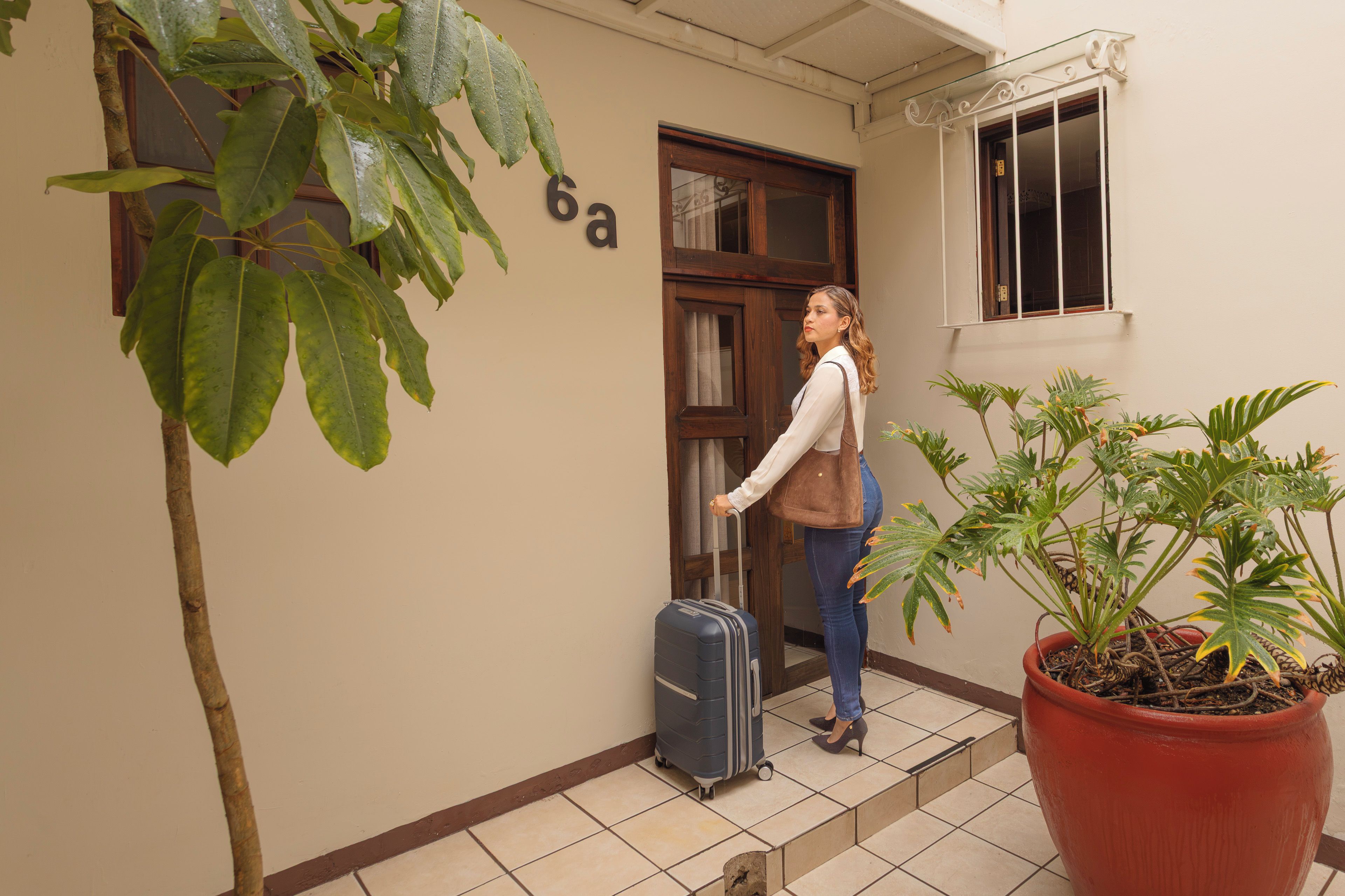 Mulher com mala parada em frente a uma porta numerada, ao lado de uma grande planta em vaso.