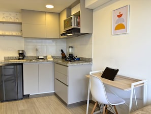Cozinha compacta com micro-ondas embutido, cafeteira e liquidificador na bancada. Pequena área de refeições com uma cadeira branca e uma mesa.