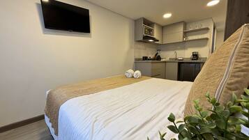 Quarto de hotel com cama, televisão de tela plana na parede, cozinha compacta com micro-ondas e cafeteira, e um pequeno vaso de planta.