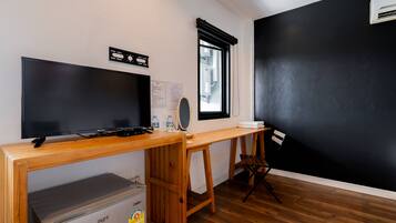 Quarto com escrivaninha de madeira, televisão de tela plana, janela com persianas, pequeno aquecedor e parede preta.