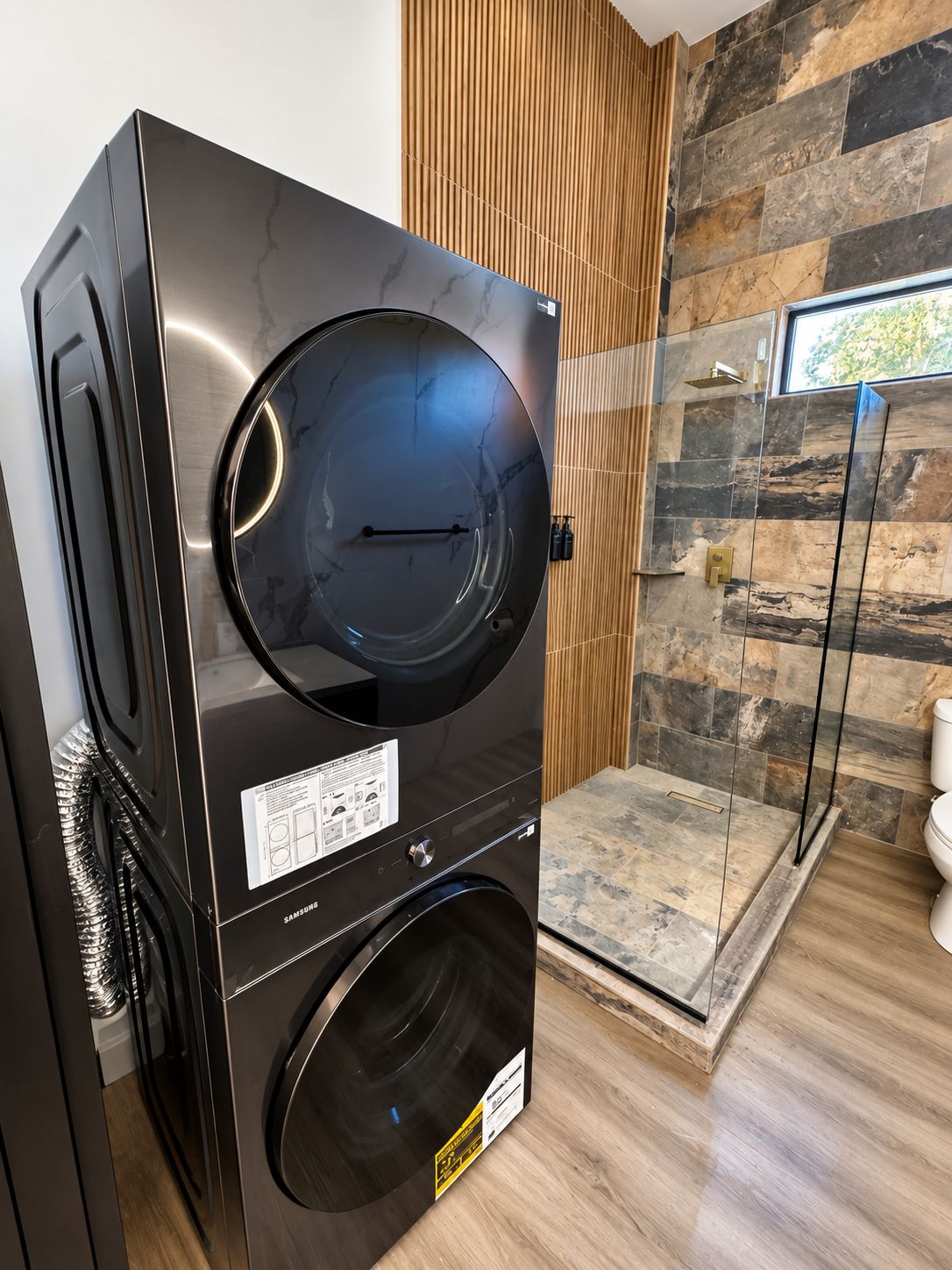 Máquina de lavar roupa e secadora pretas num banheiro moderno com parede de pedra e box de vidro.