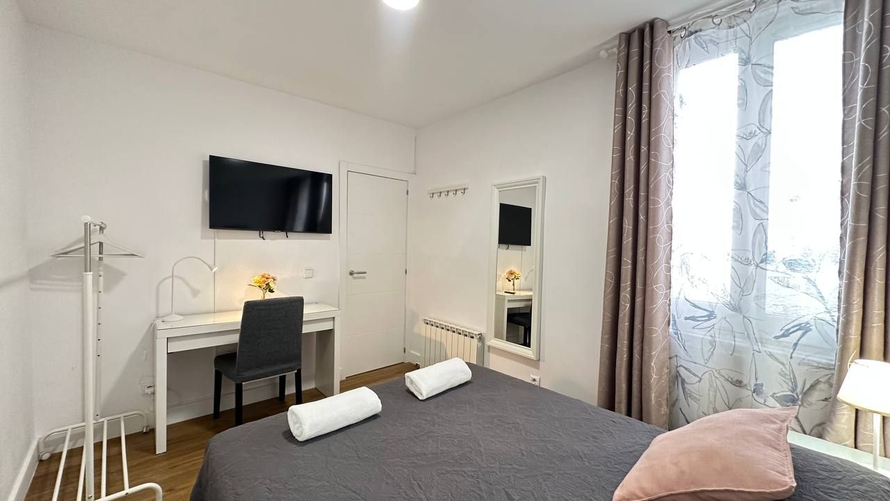 Quarto com cama, escrivaninha, cadeira, televisão, espelho e janela com cortinas.