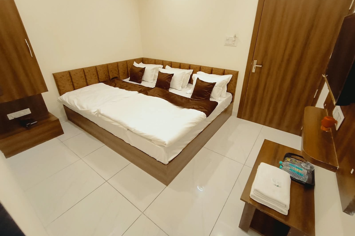 Cama bem arrumada em quarto de hotel, com cabeceira de madeira, mesa de cabeceira com abajur e uma pequena mesa com toalha e bolsa.