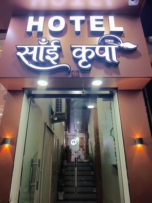 Entrada de hotel com uma placa em inglês e outro idioma, escadaria que leva para o interior e um pequeno santuário com um adorno pendurado.