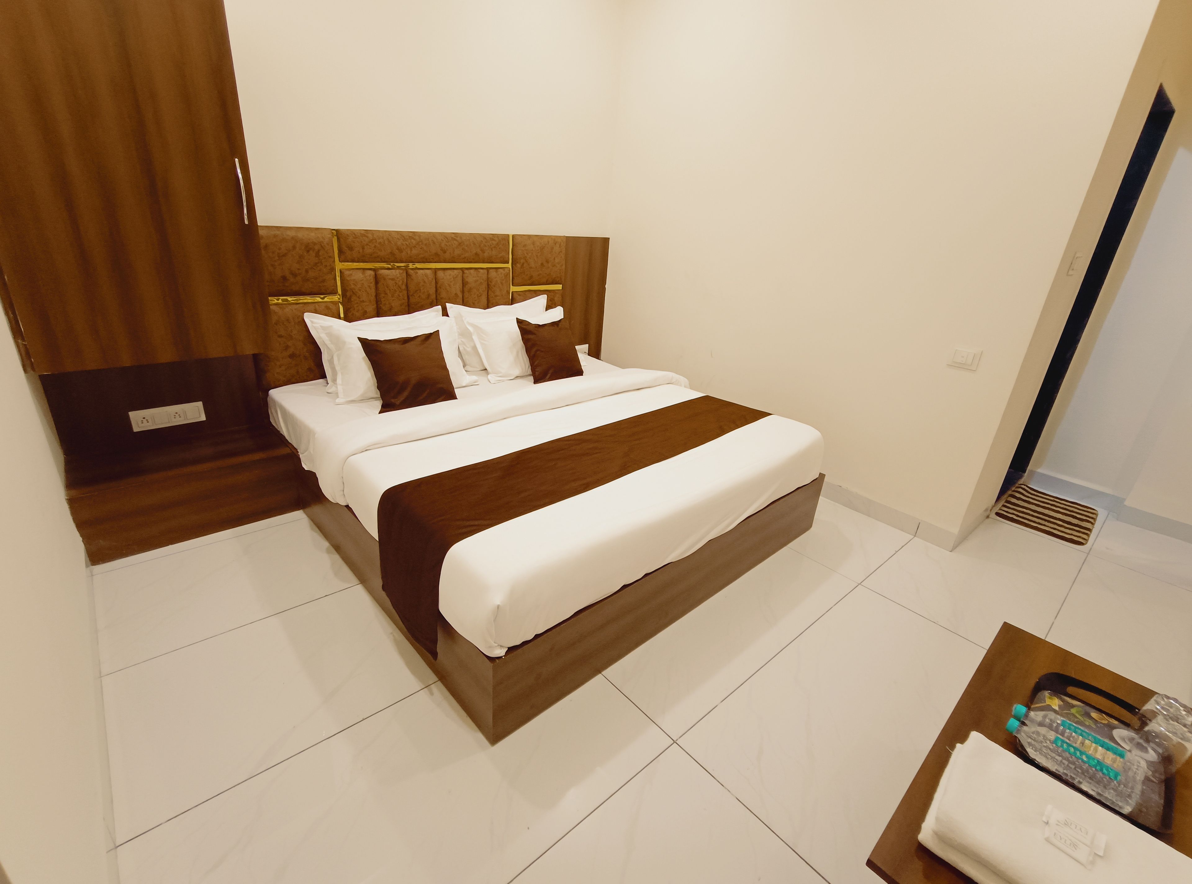 Quarto de hotel com uma cama grande, cabeceira de madeira, mesa de cabeceira com uma garrafa e lenços, e uma tomada na parede.