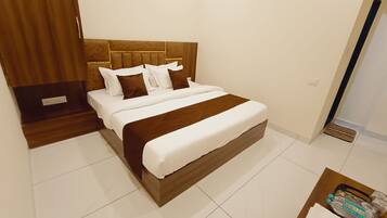 Quarto de hotel com uma cama grande, cabeceira de madeira, mesa de cabeceira com uma garrafa e lenços, e uma tomada na parede.