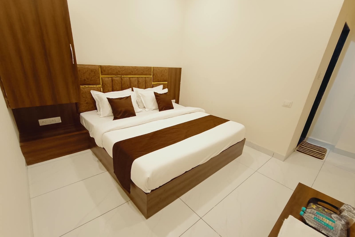 Quarto de hotel com cama, cabeceira de madeira, roupa de cama branca e uma pequena mesa com uma bolsa e uma caixa.