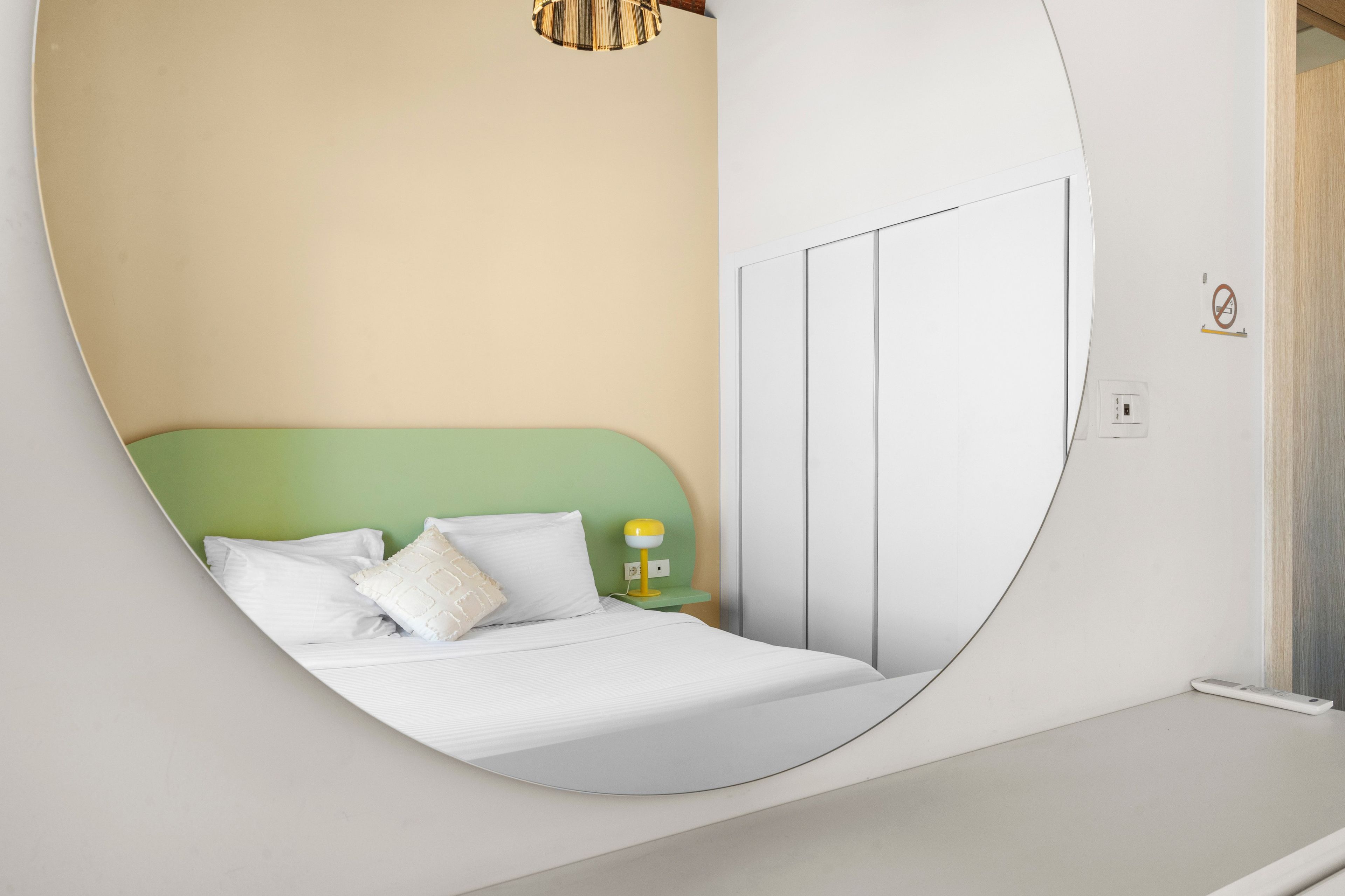 Quarto com espelho curvo, cama com roupa de cama branca, cabeceira verde e mesa de cabeceira com abajur.