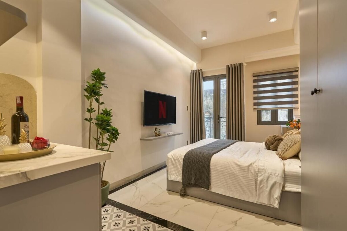Quarto moderno com cama, televisão fixada na parede, janela com cortinas e piso de mármore.