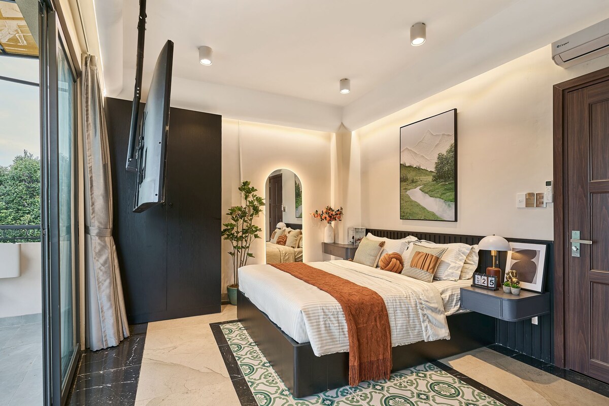 Quarto moderno com uma cama grande, mesa de cabeceira com abajur, uma obra de arte na parede e uma porta com fechadura digital.