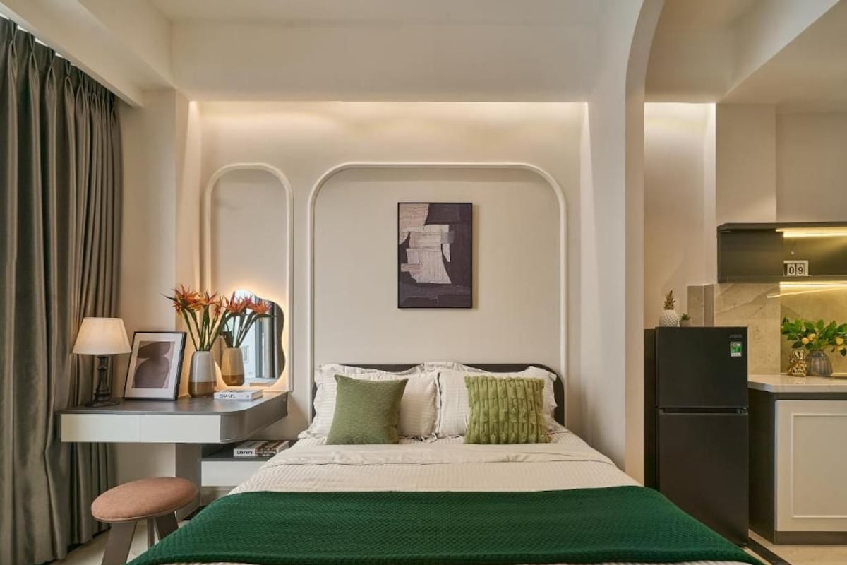 Quarto de hotel com cama, escrivaninha com luminária, cadeira, pequena mesa com flores e cozinha equipada com geladeira.