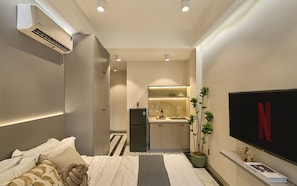 Quarto de hotel compacto com uma cama, pequeno espaço de cozinha, TV e planta em vaso.