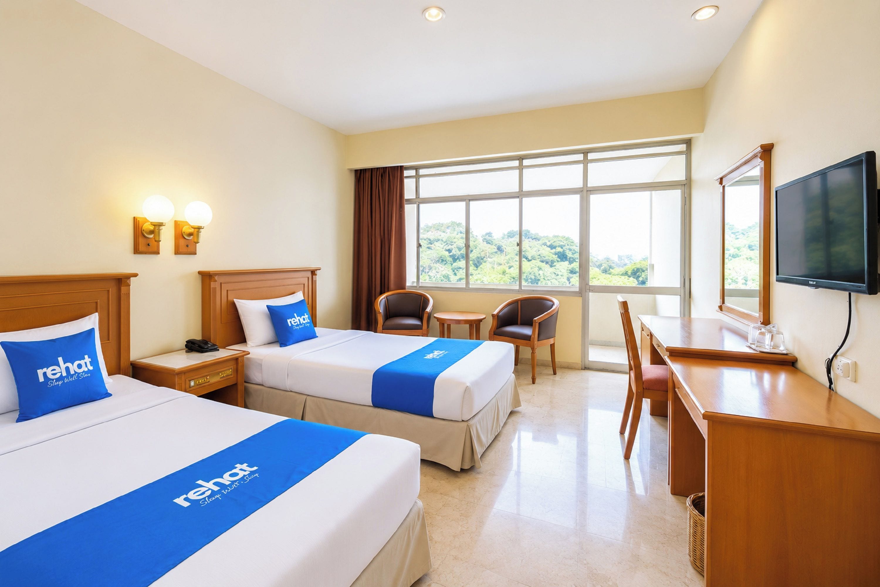 Quarto de hotel com duas camas, uma escrivaninha, uma cadeira, uma televisão e janela com vista para árvores.