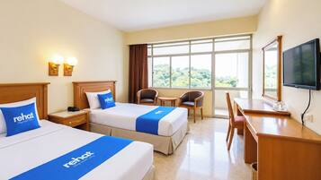 Quarto de hotel com duas camas, uma escrivaninha, uma cadeira, uma televisão e janela com vista para árvores.
