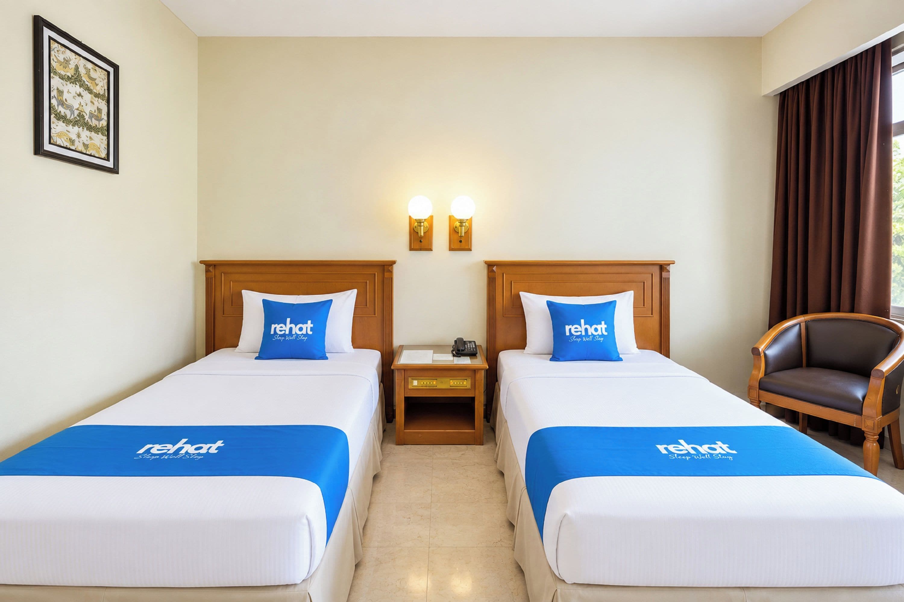Quarto de hotel com duas camas de solteiro, cada uma com colcha azul e travesseiro com o logo 'rehot'. Há duas luminárias fixadas na parede, uma mesa de cabeceira de madeira com um telefone e uma cadeira.