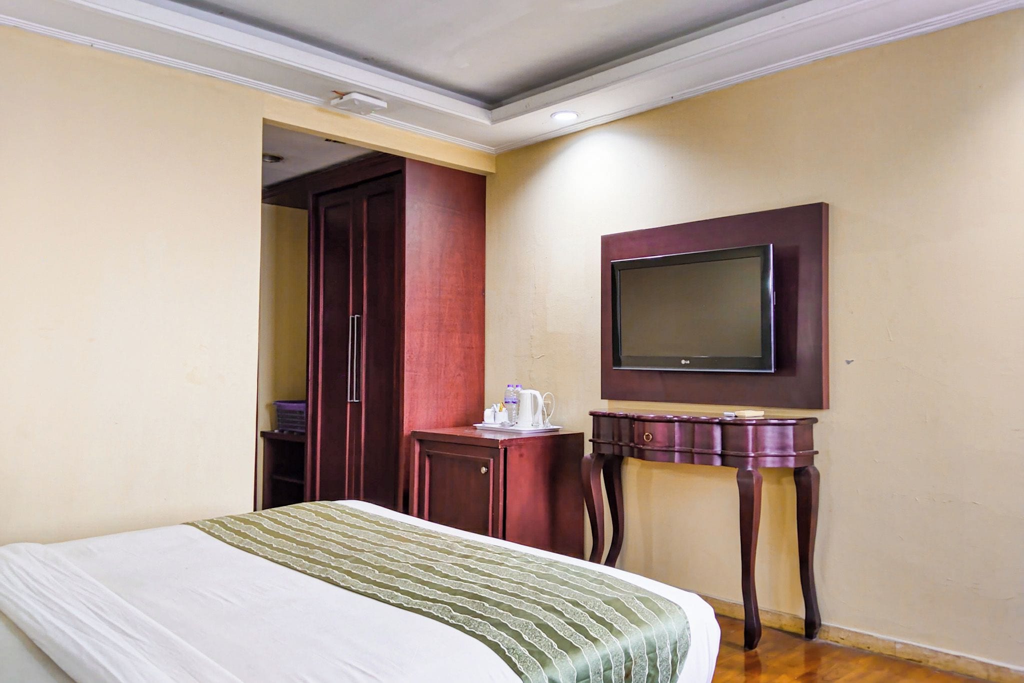 Quarto de hotel com uma cama, televisão fixada na parede, um armário de madeira e uma porta que dá para outro cômodo.