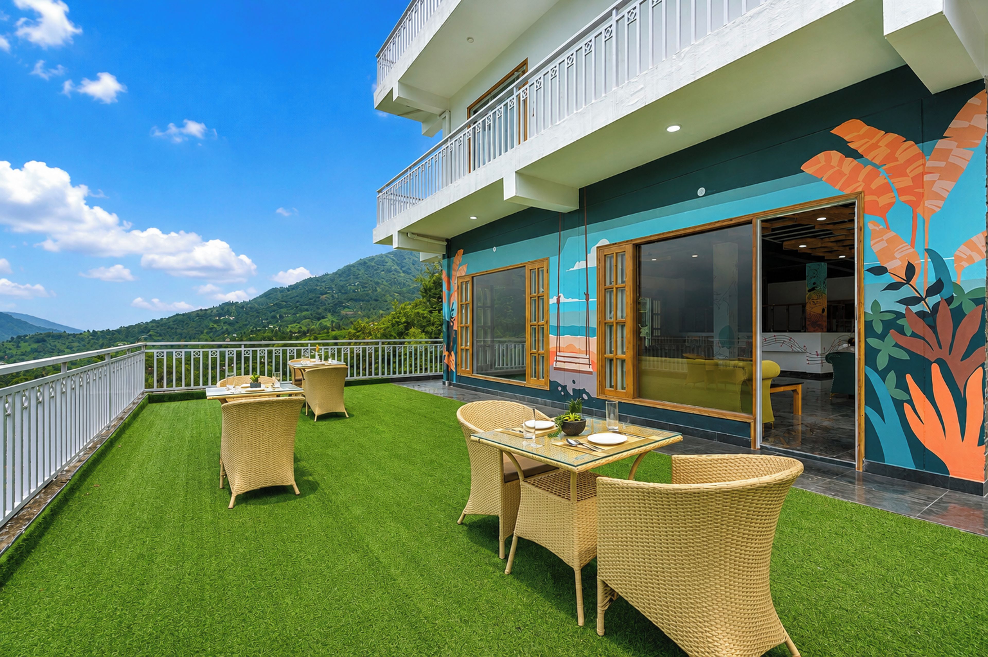 Varanda de hotel com grama artificial, móveis de vime e vista para as montanhas.