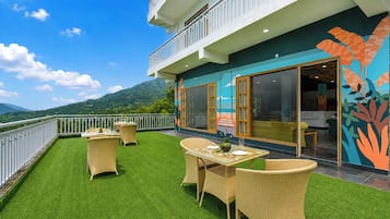 Varanda de hotel com grama artificial, móveis de vime e vista para as montanhas.