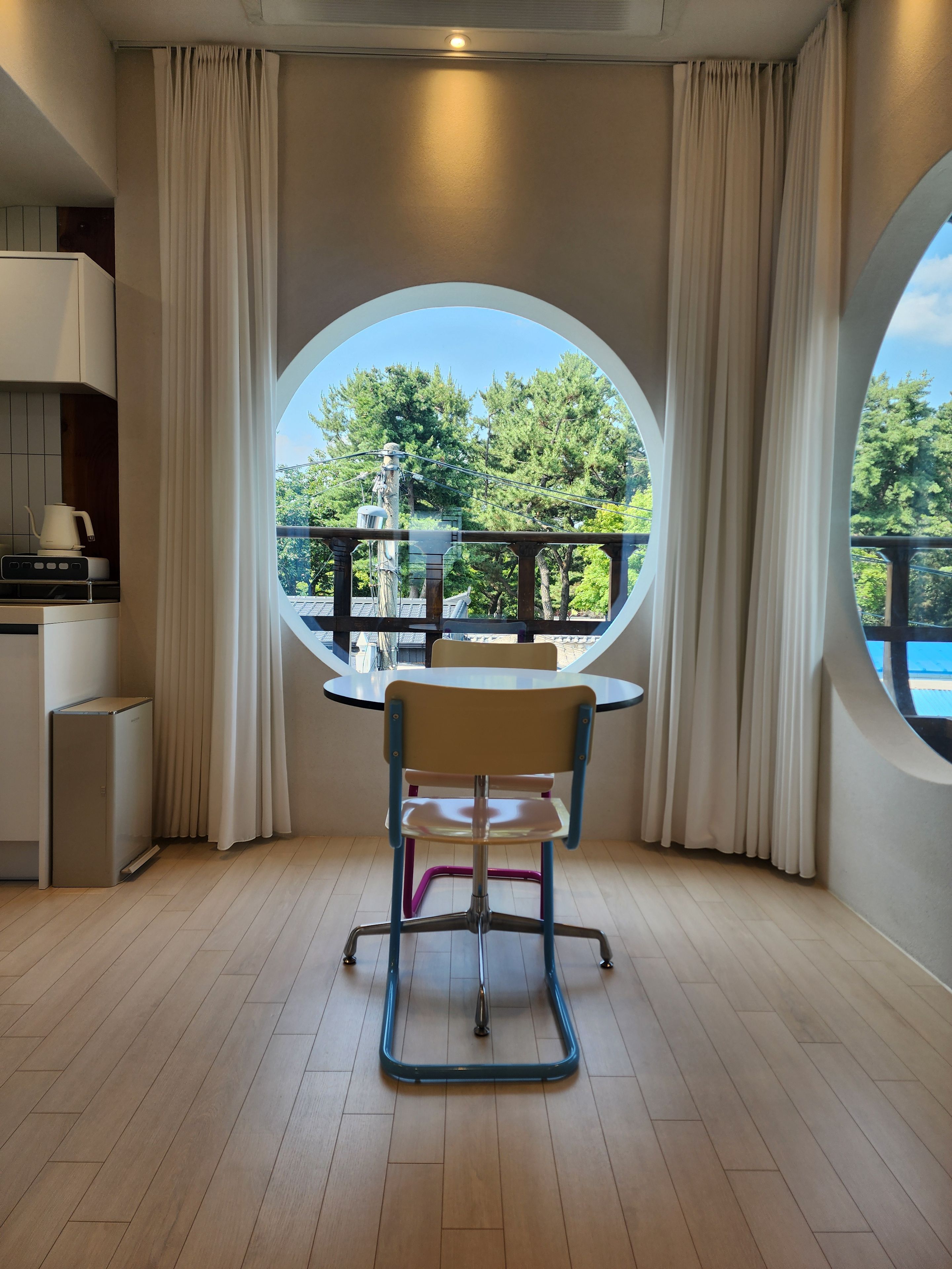 Uma cozinha moderna com uma mesa redonda e uma cadeira, vista para árvores e uma varanda através de uma janela circular, e uma luminária no teto.
