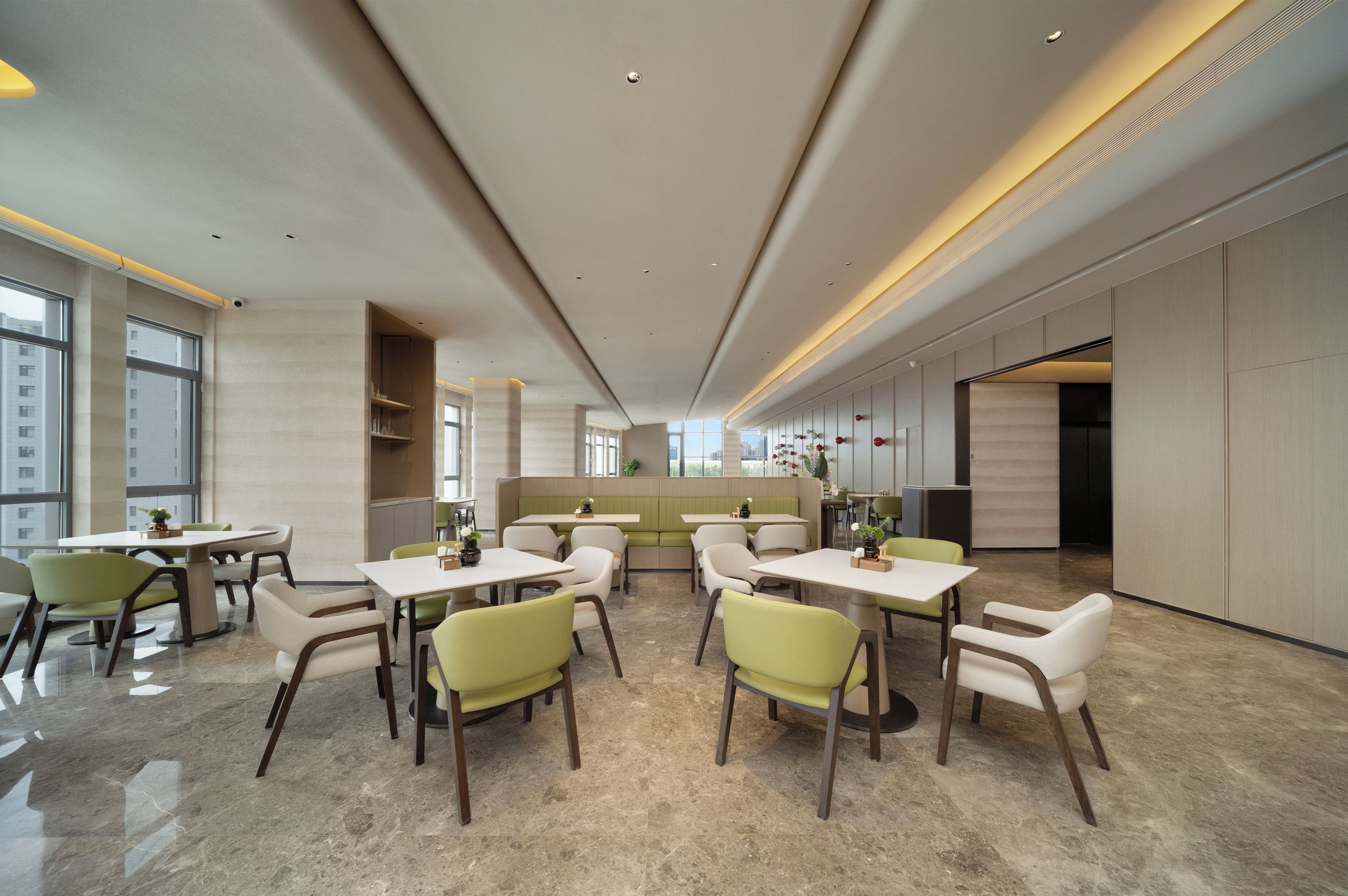 Uma sala de jantar moderna com várias mesas e cadeiras, teto alto com iluminação embutida e grandes janelas que oferecem luz natural.