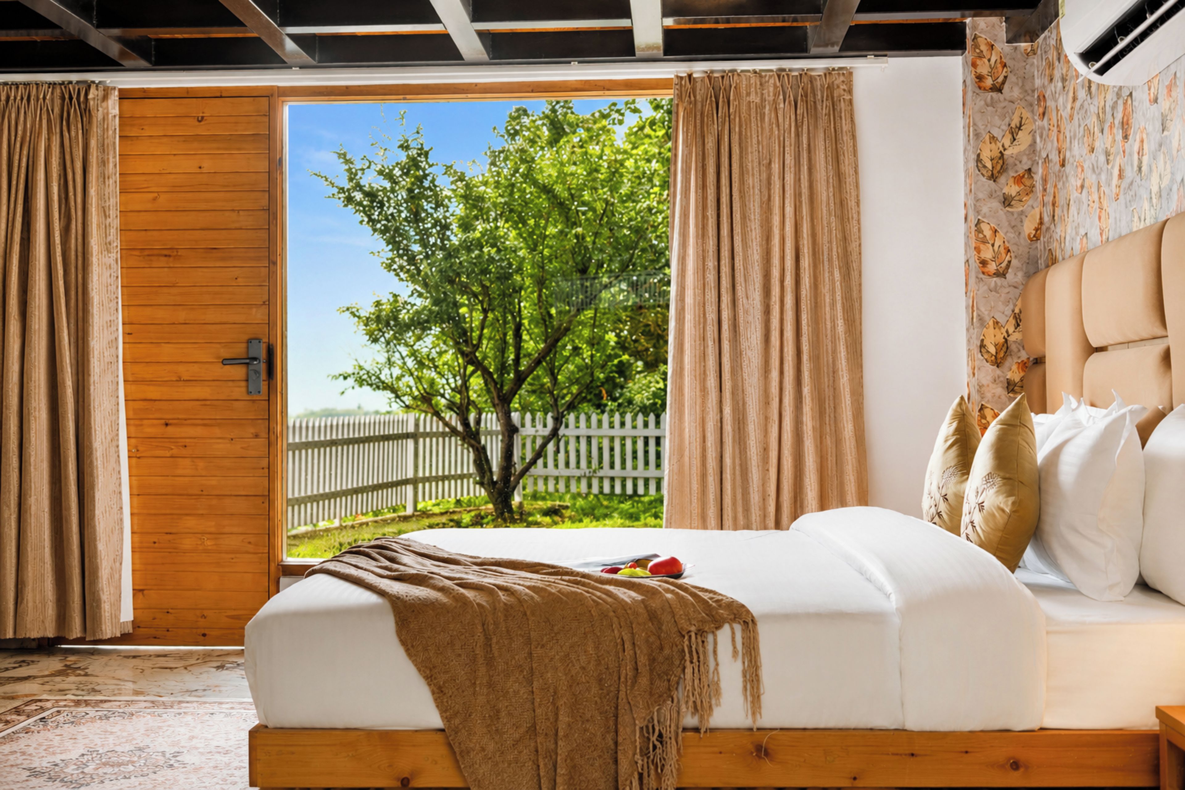 Quarto com cama, cabeceira de madeira, cortinas e vista para uma árvore e cerca.
