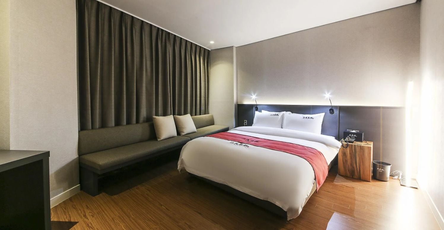 Quarto de hotel moderno com cama, sofá, mesa de cabeceira e abajur.