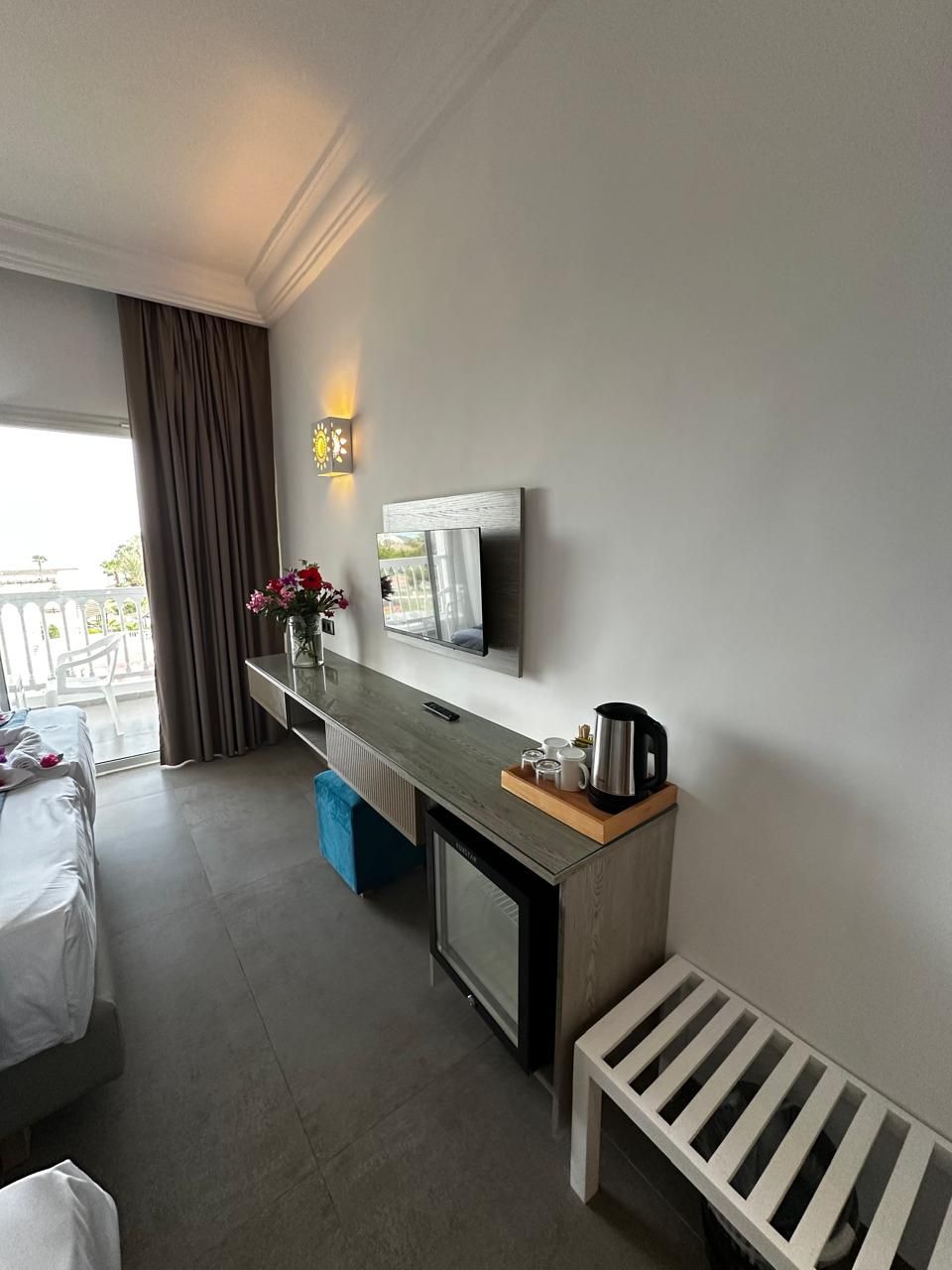 Quarto de hotel com cozinha compacta, cama, mesa pequena com um vaso de flores e vista para o exterior.