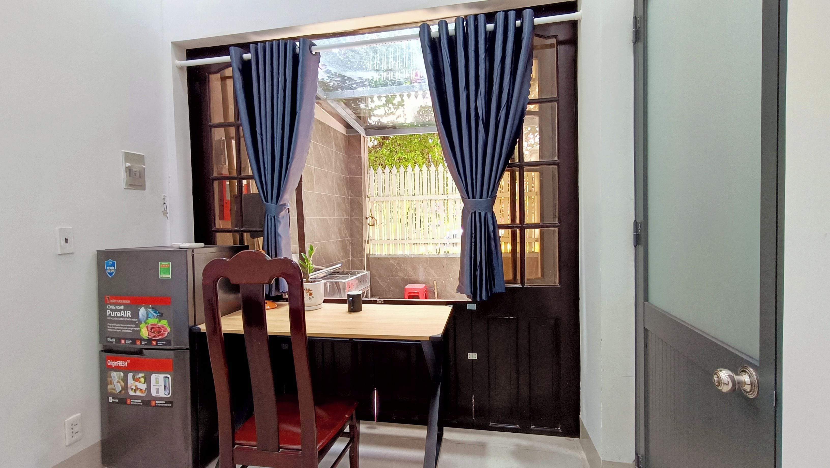 Quarto compacto com uma mesa de madeira, uma cadeira, um pequeno frigorífico e janela com cortinas.