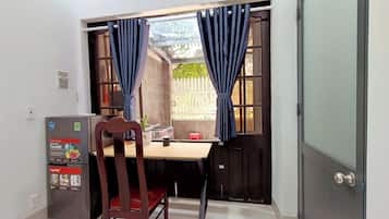 Quarto compacto com uma mesa de madeira, uma cadeira, um pequeno frigorífico e janela com cortinas.