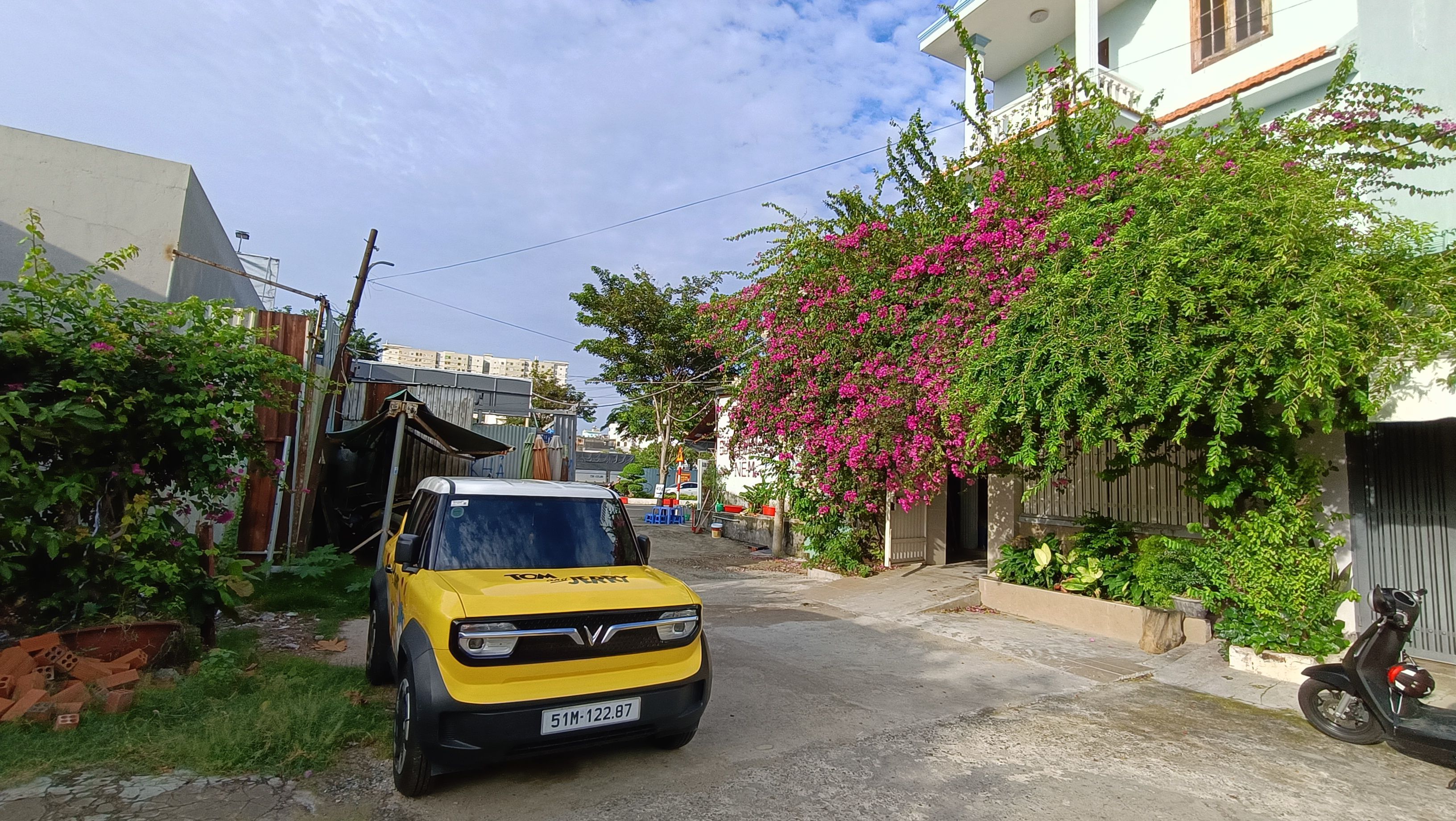 Rua estreita com um veículo amarelo estacionado na lateral, cercada por vegetação e edifícios.