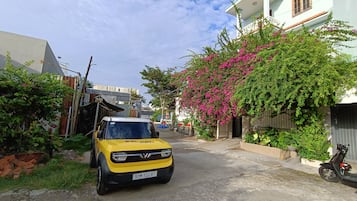 Rua estreita com um veículo amarelo estacionado na lateral, cercada por vegetação e edifícios.