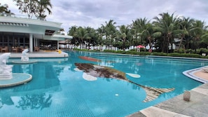 Piscina com escultura de golfinho, palmeiras e um edifício com área de estar ao ar livre.