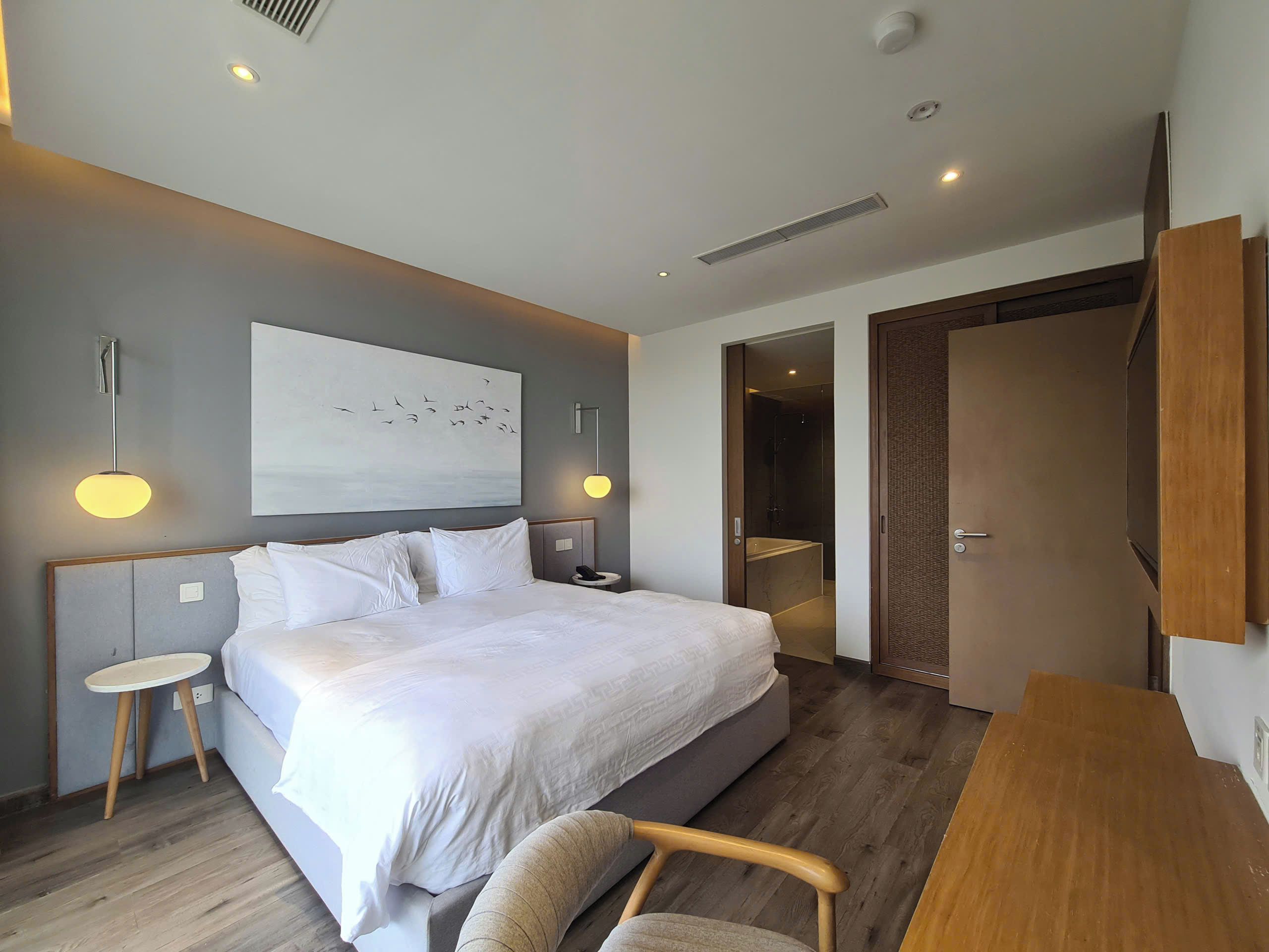 Quarto de hotel moderno com uma cama grande, mesas de cabeceira, escrivaninha e banheiro visível através de uma porta aberta.