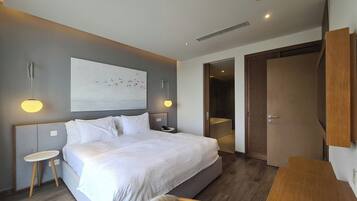 Quarto de hotel moderno com uma cama grande, mesas de cabeceira, escrivaninha e banheiro visível através de uma porta aberta.
