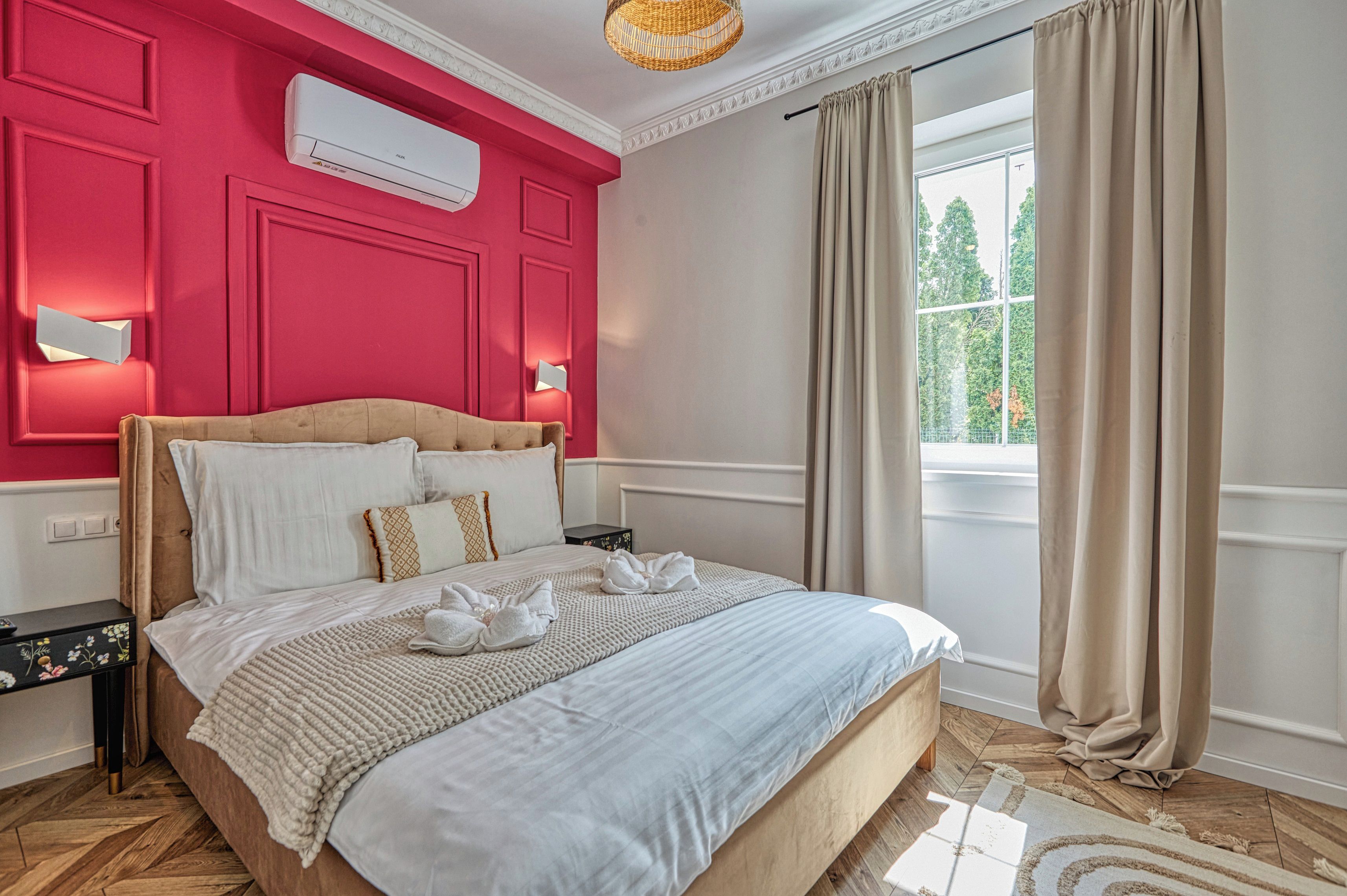 Quarto de hotel com uma cama grande, uma parede de destaque vermelha, uma janela com cortinas e um ar condicionado fixado na parede.