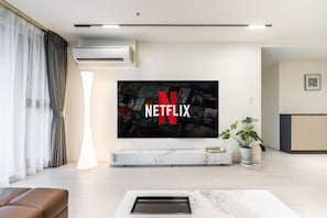Uma sala de estar moderna com uma grande televisão de tela plana na parede, um console branco, um sofá de couro marrom e um abajur de chão alto.