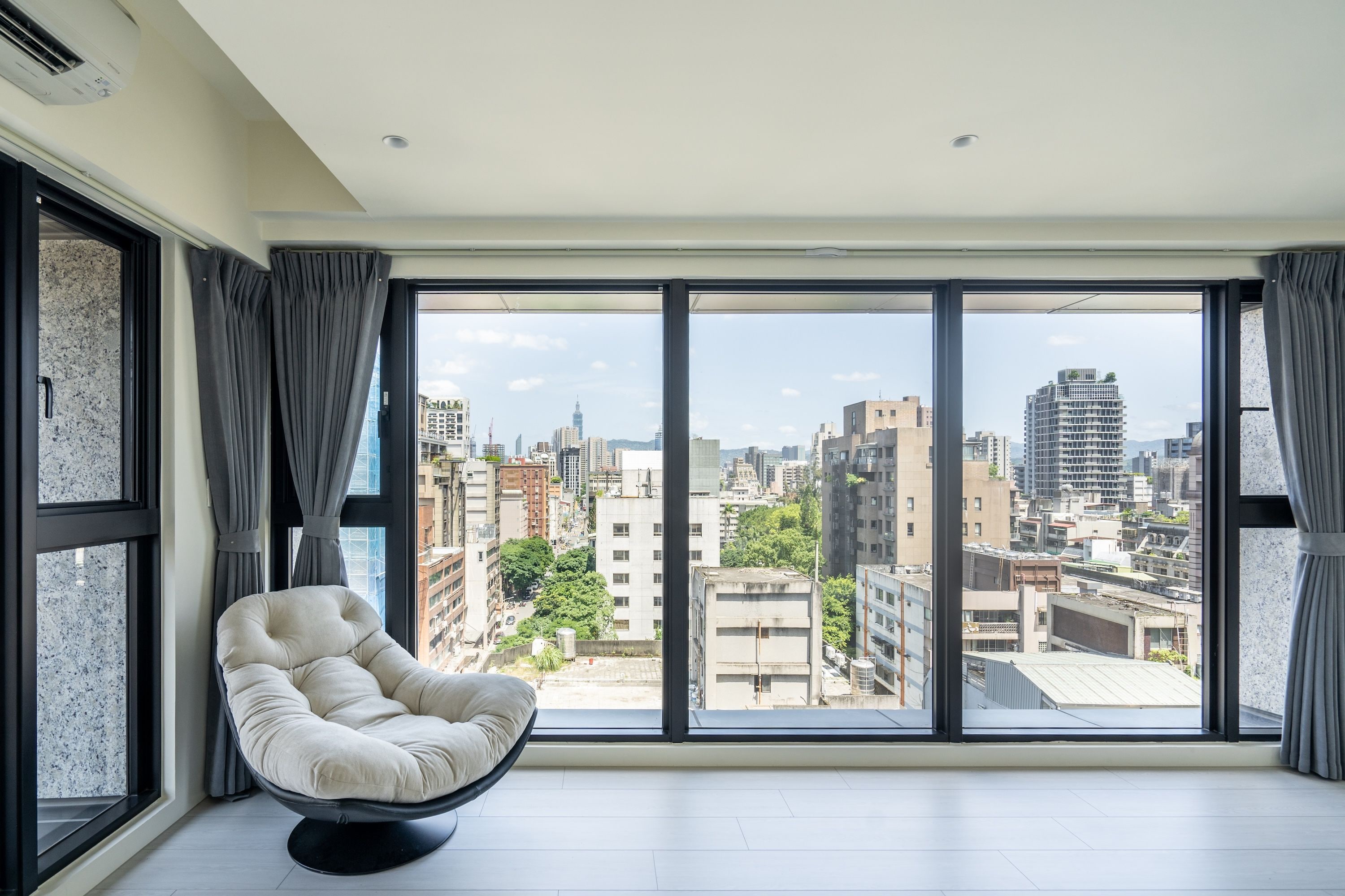 Quarto moderno com uma grande janela, uma poltrona estofada e vista para a paisagem urbana.