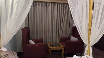 Quarto com duas poltronas, uma mesinha e uma cama com roupa de cama branca.